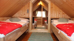 Um quarto de sótão com piso de madeira e duas camas, cada uma com um cobertor vermelho e um travesseiro xadrez. Um tapete verde está no chão, e uma escada leva a um mezanino com uma janela.