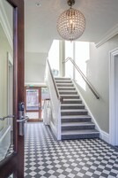 Uma escadaria com piso xadrez que leva a um patamar, um lustre e uma porta com dispositivo de segurança.