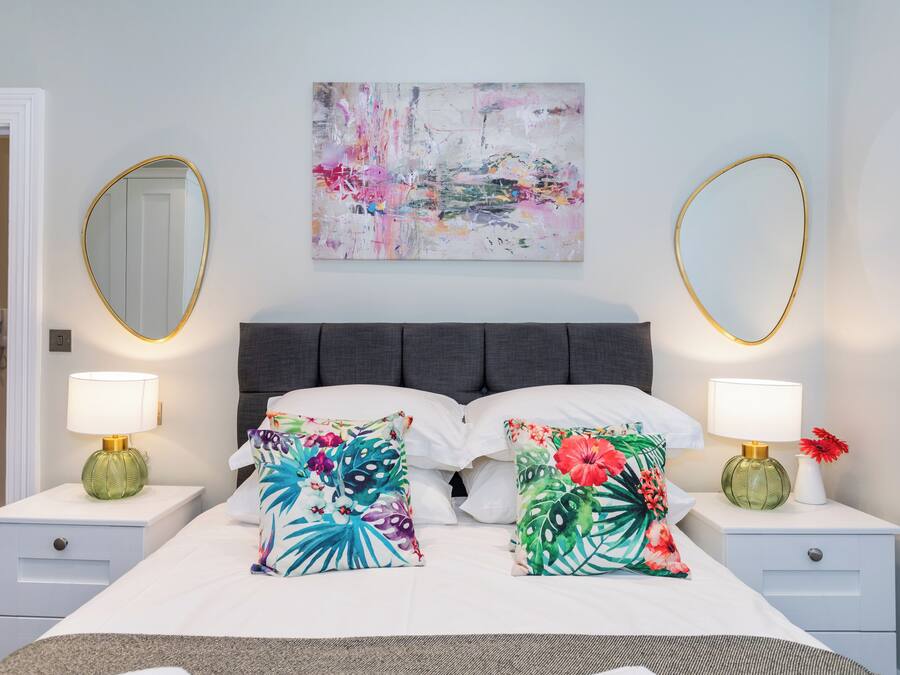 Cama bem arrumada com almofadas florais, duas luminárias de cabeceira e um quadro abstrato grande na parede.