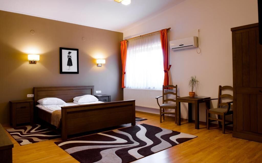 Quarto com cama, mesas de cabeceira, escrivaninha, cadeiras, radiador e janela com cortinas.
