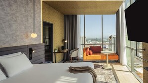 Quarto de hotel moderno com uma cama grande, uma escrivaninha, uma cadeira e um sofá. Há uma TV fixada na parede e uma janela com vista para a cidade.