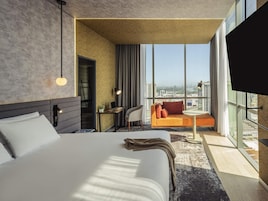 Quarto de hotel moderno com uma cama grande, uma escrivaninha, uma cadeira e um sofá. Há uma TV fixada na parede e uma janela com vista para a cidade.