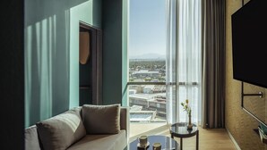 Quarto de hotel moderno com sofá, uma mesinha com chávenas, televisão e uma janela ampla com vista para a cidade.