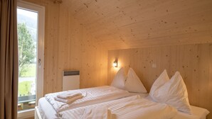 Cama de casal com lençóis e almofadas brancas, mesa de cabeceira, luminária fixada na parede e janela com cortinas.
