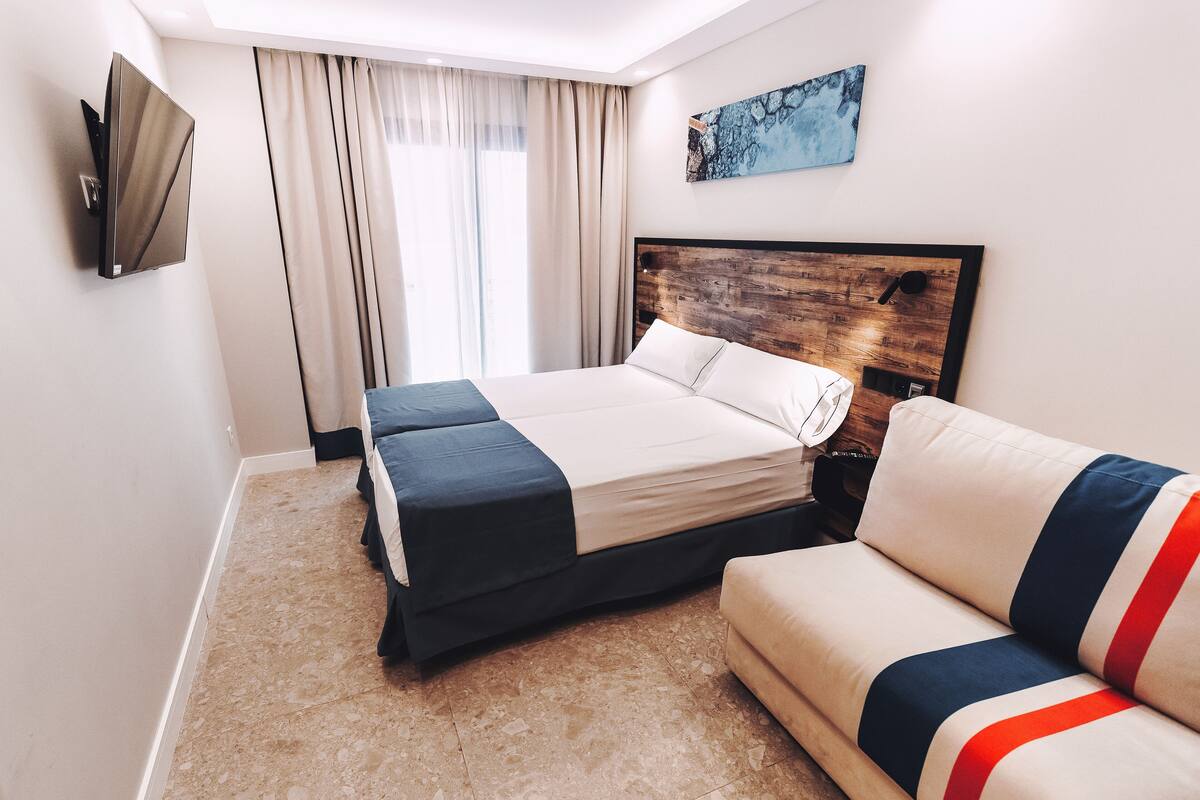Quarto de hotel com cama, sofá, televisão de tela plana, quadro e janela com cortinas.