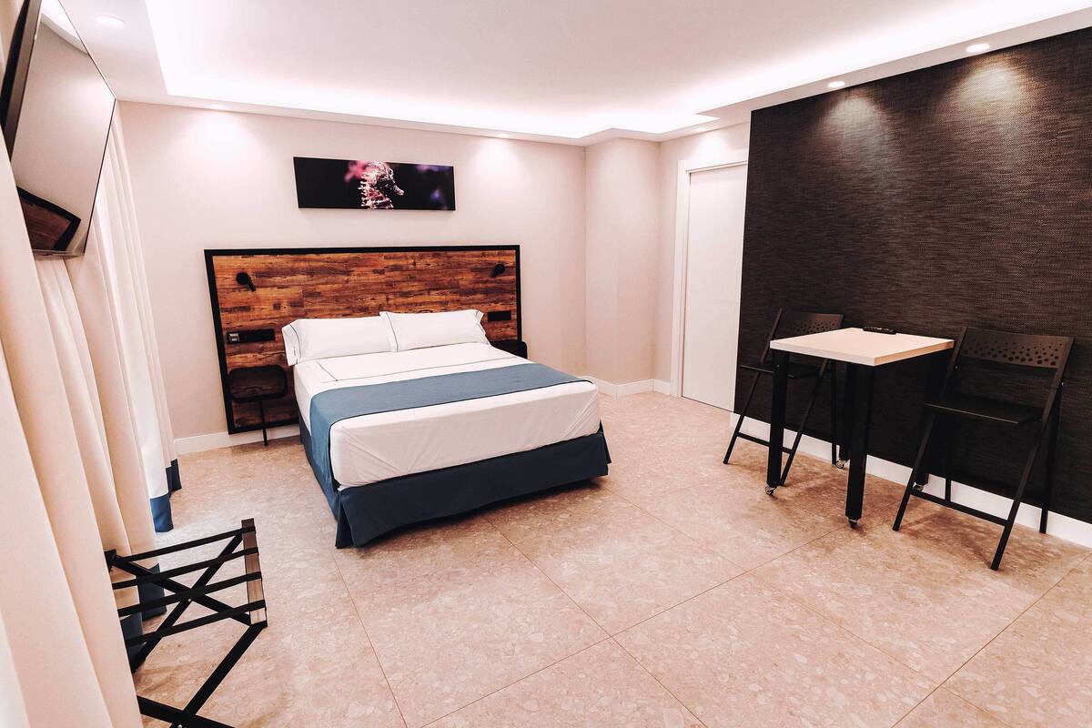 Quarto moderno com cama de cabeceira em madeira, uma mesinha com cadeiras e uma TV na parede.
