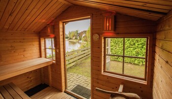 Um salão de sauna de madeira com um banco, uma porta que dá para o exterior e uma janela com vista para um jardim e um corpo d'água.