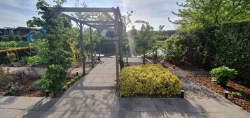 Um caminho de jardim com pérgola de madeira, cercado por arbustos e árvores diversas.