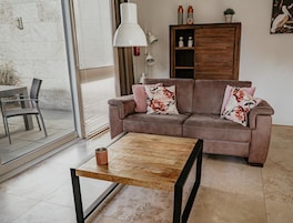 Sala de estar com um sofá marrom, duas almofadas florais, uma mesa de centro de madeira e uma porta de vidro que dá para o exterior.