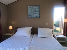 Uma cama de casal com roupa de cama branca, duas mesas de cabeceira com luminárias, um letreiro fixado na parede e uma janela com vista para o verde.