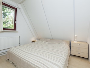 Quarto com teto inclinado, janela com vista para árvores, radiador, mesa de cabeceira com abajur e cama com manta listrada.