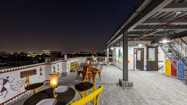 Terraço no telhado com mesas, cadeiras, um candeeiro e vista da cidade à noite.