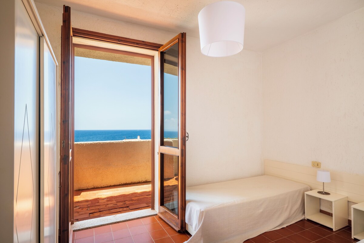 Quarto com cama, mesa de cabeceira e vista para o mar através de uma porta aberta.