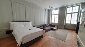 Quarto com cama grande, sofá, lustre e janelas amplas.