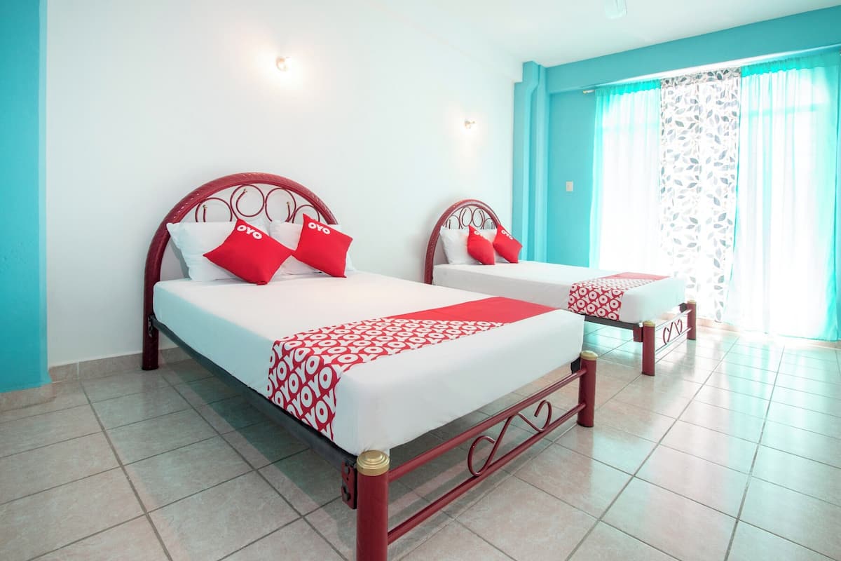 Duas camas com roupa de cama vermelha e branca, uma parede azul e uma janela com cortinas finas.
