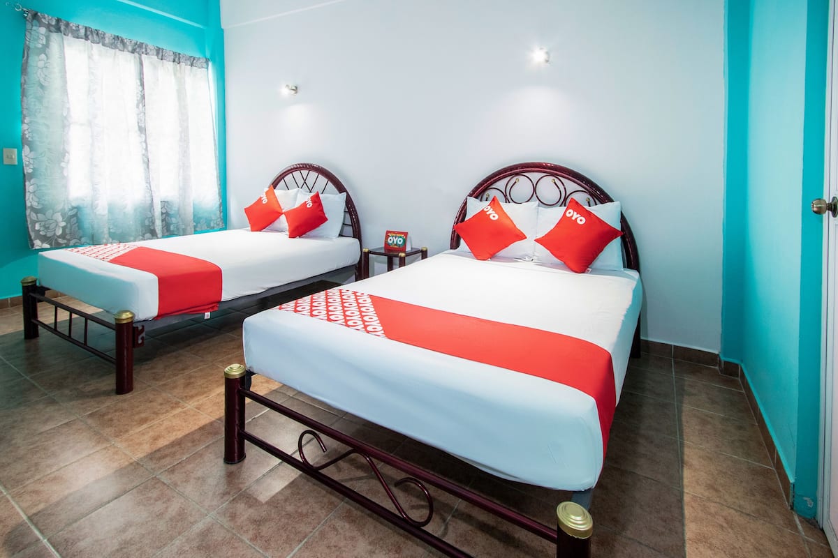 Duas camas de solteiro com roupa de cama vermelha e branca, uma mesinha com um relógio e uma janela com cortinas florais.