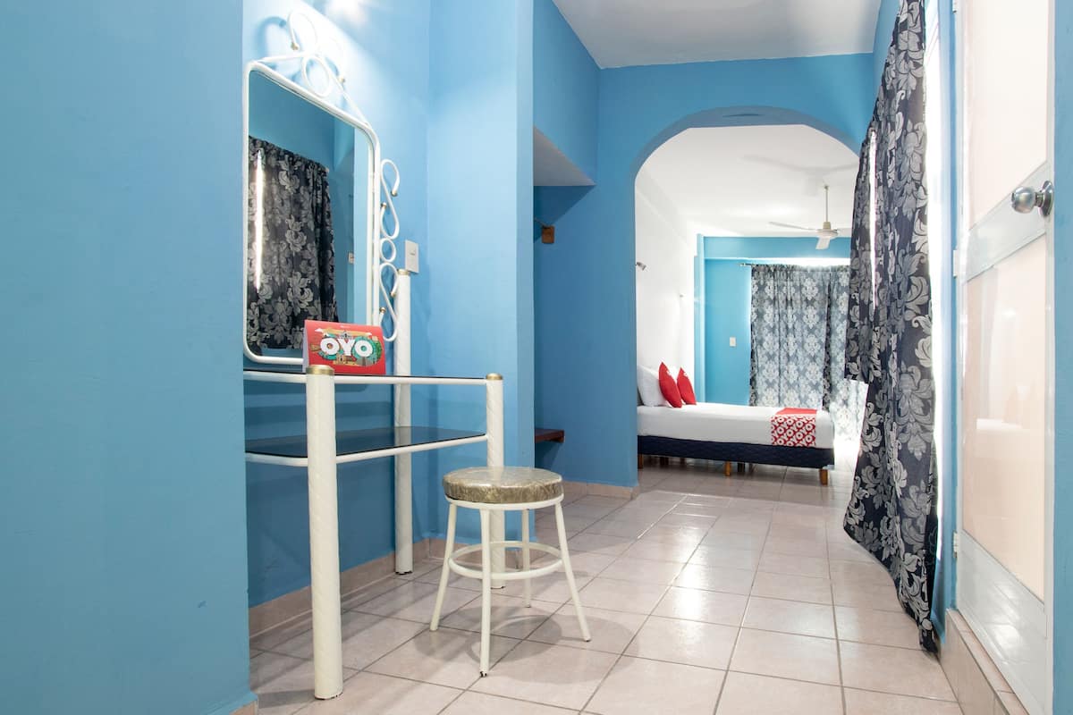 Quarto com paredes azuis, cama com cabeceira branca, um banquinho pequeno e espelho com moldura decorativa.