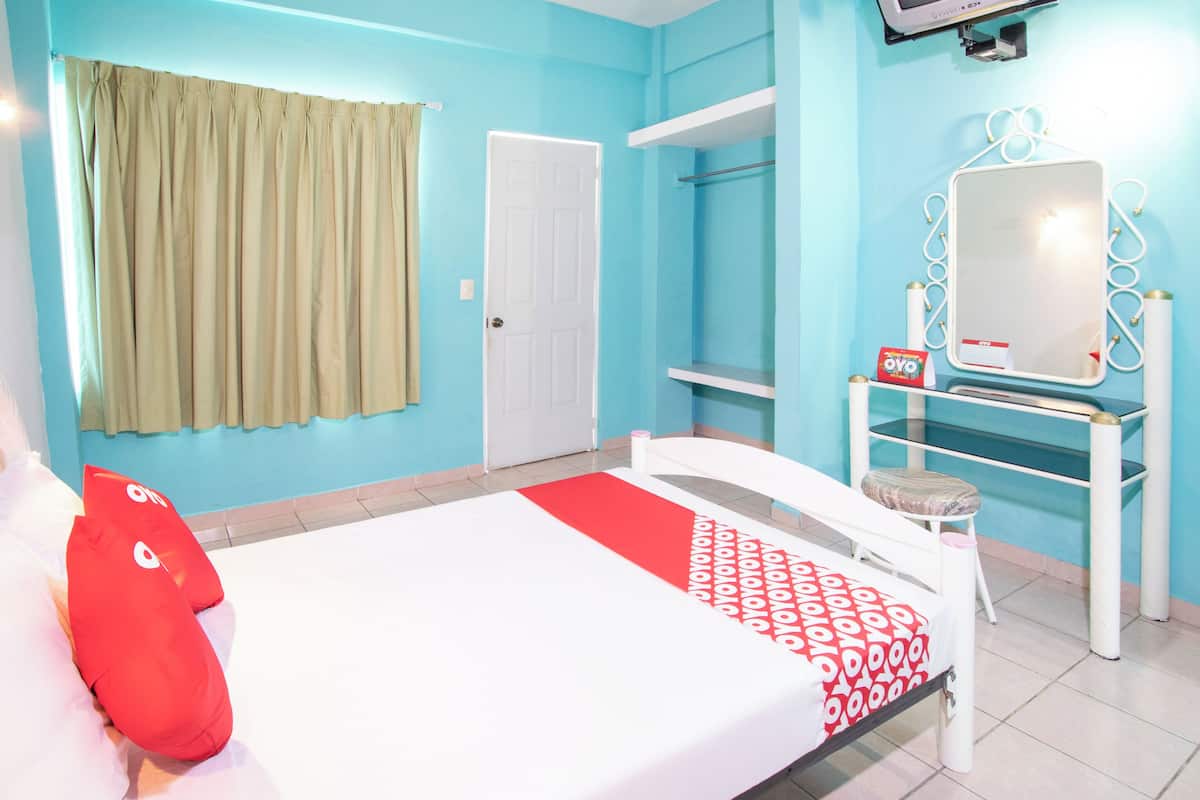 Quarto de hotel com cama, toucador com espelho, uma porta e uma prateleira com um objeto vermelho.