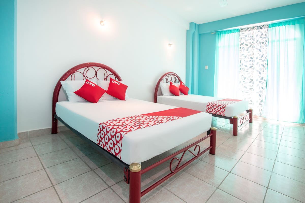 Quarto de hotel com duas camas, cada uma com colcha e almofadas estampadas em vermelho e branco. O quarto possui piso de azulejo e janela com cortina azul.