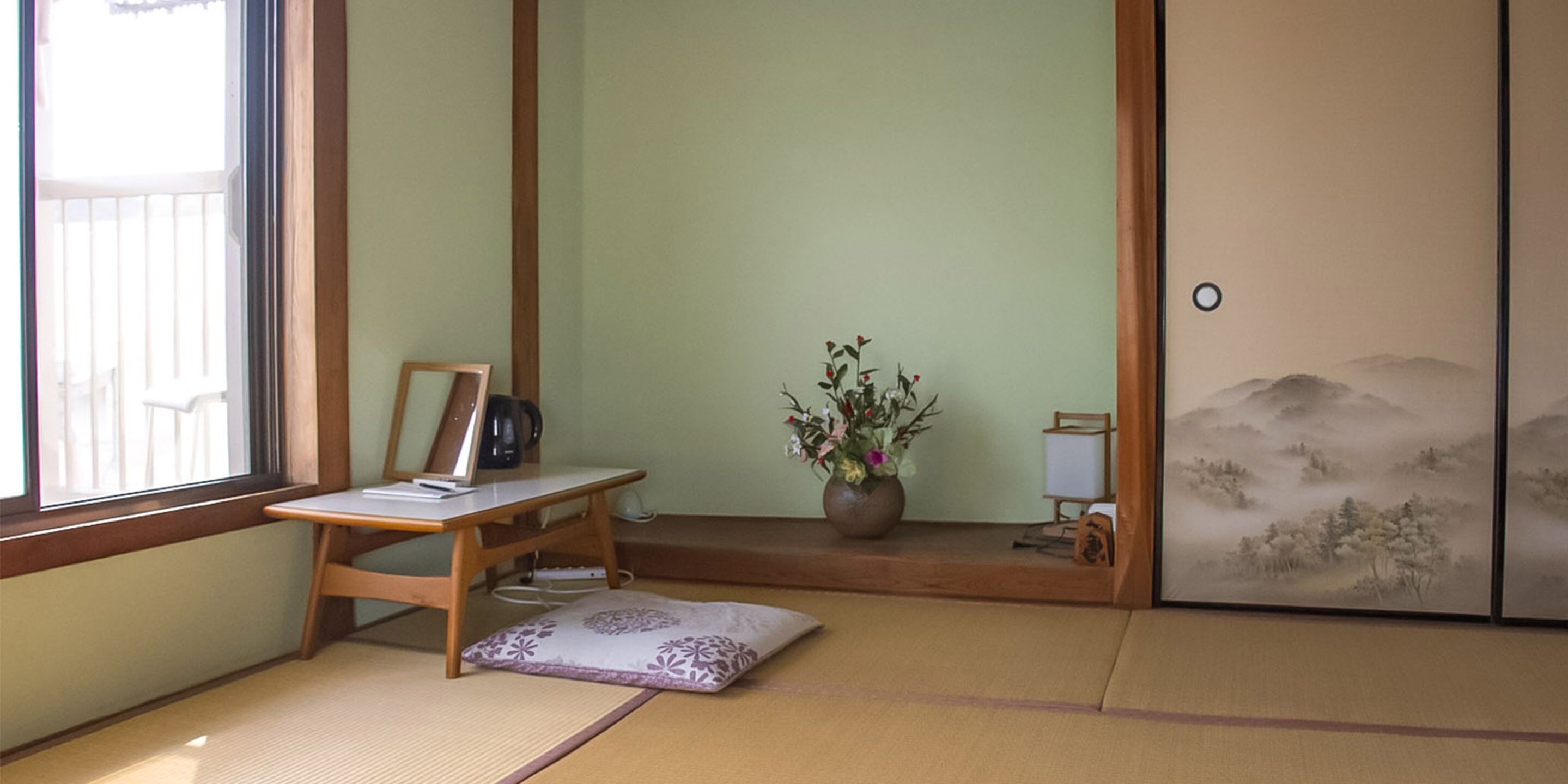 Um quarto japonês tradicional, com piso de tatame, uma mesa baixa com um porta-retrato, um vaso com flores e um biombô com pintura de paisagem.