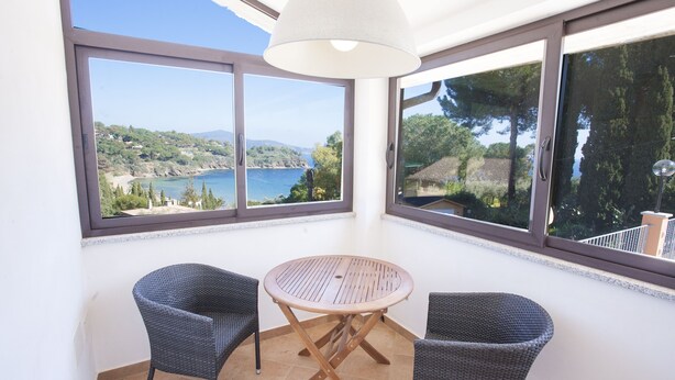 Quarto de canto com duas cadeiras de vime, uma mesa de madeira e grandes janelas com vista para o mar e colinas.