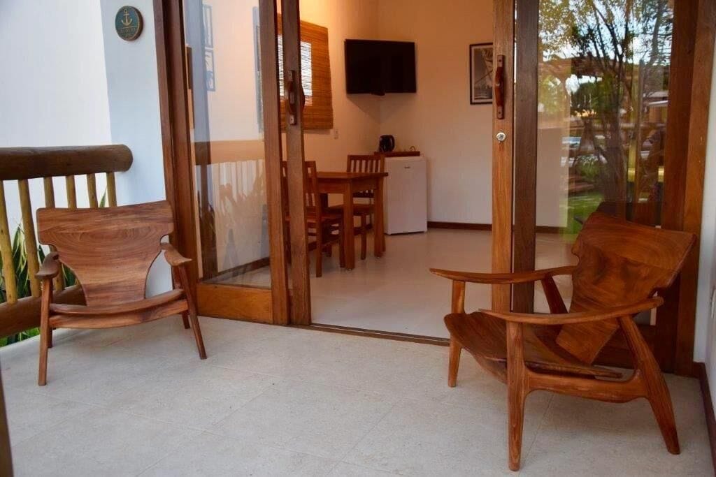 Uma cadeira de madeira e uma mesa numa varanda, com vista para uma área de refeições e uma televisão na parede.