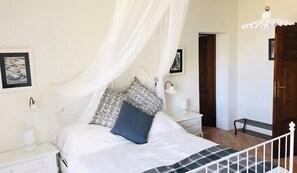 Quarto com cama sob dossel, mesas de cabeceira, mesa de apoio, abajur e porta.