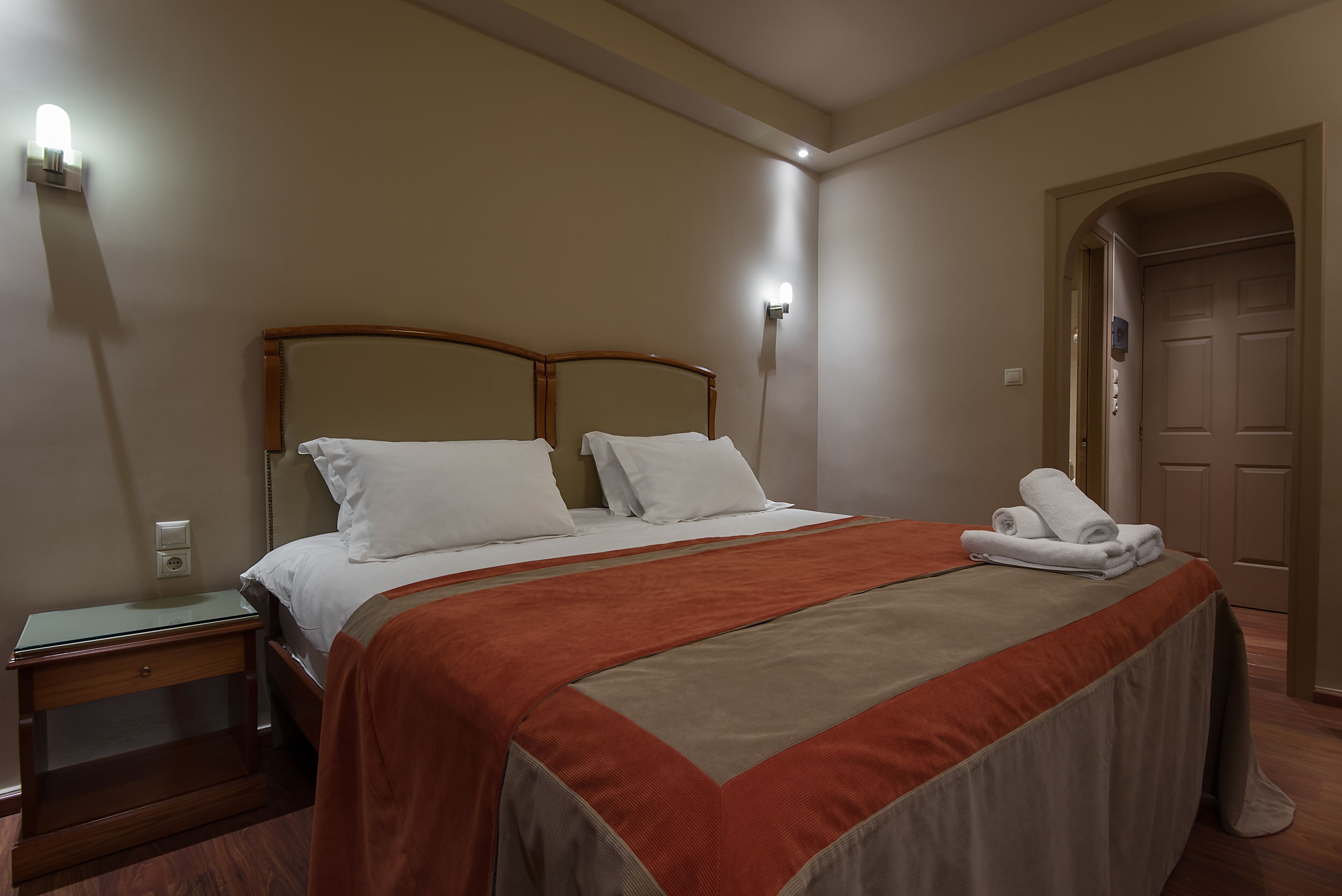 Quarto de hotel com cama, mesa de cabeceira, luminária na parede e porta.
