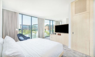 Quarto de hotel com uma cama grande, televisão, cadeira e vista para um edifício e um espelho d'água.