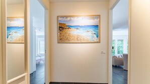 Um corredor com um quadro de uma praia na parede, uma porta e vista para um quarto com cama e janela.