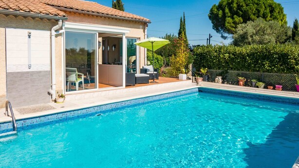 Uma piscina com borda azulejada, um alpendre coberto com mobiliário de exterior e um edifício com telhado e portas de vidro.