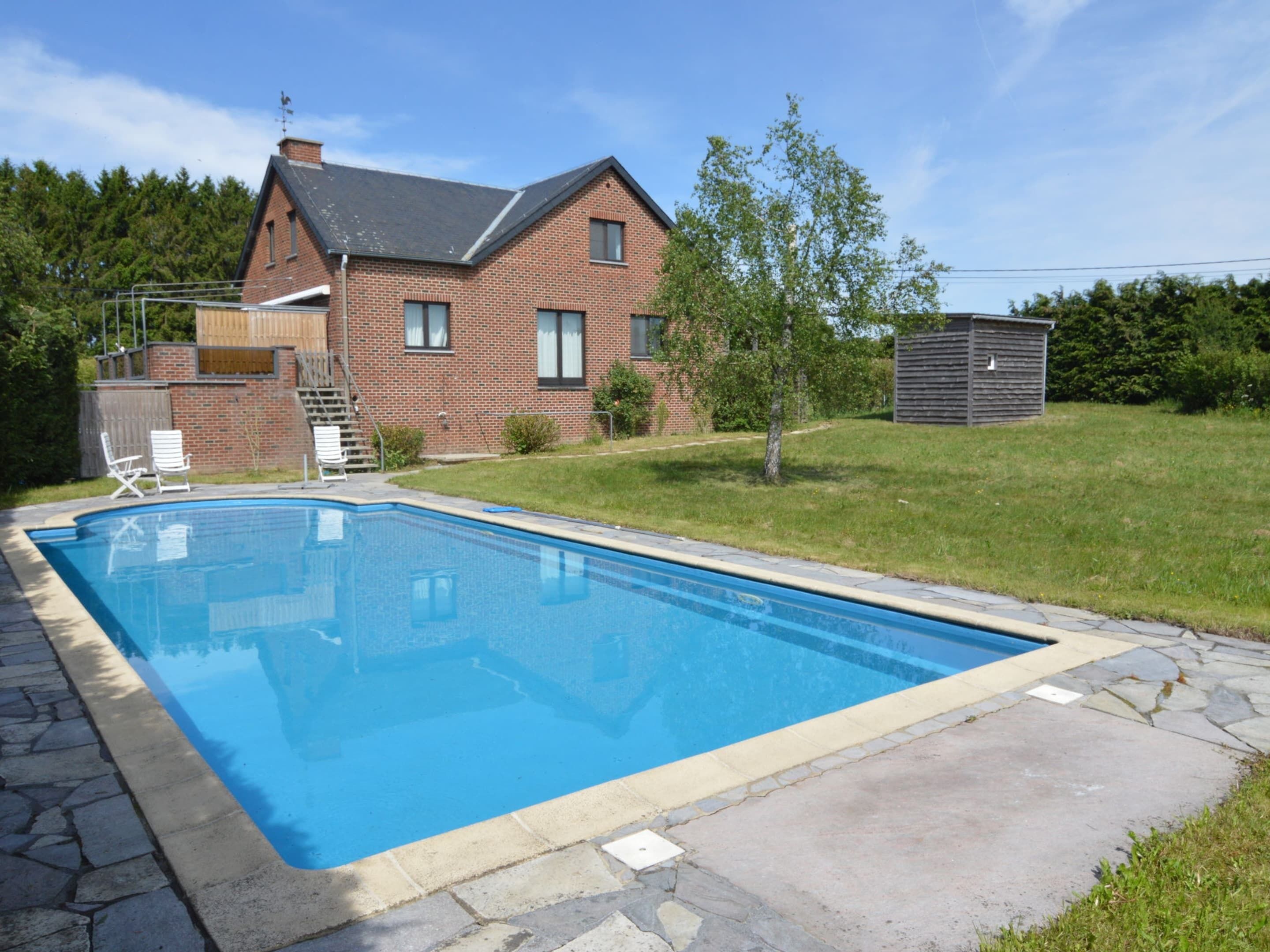 Uma piscina retangular com borda de pedra, cercada por um gramado, uma casa de tijolos e um pequeno galpão.