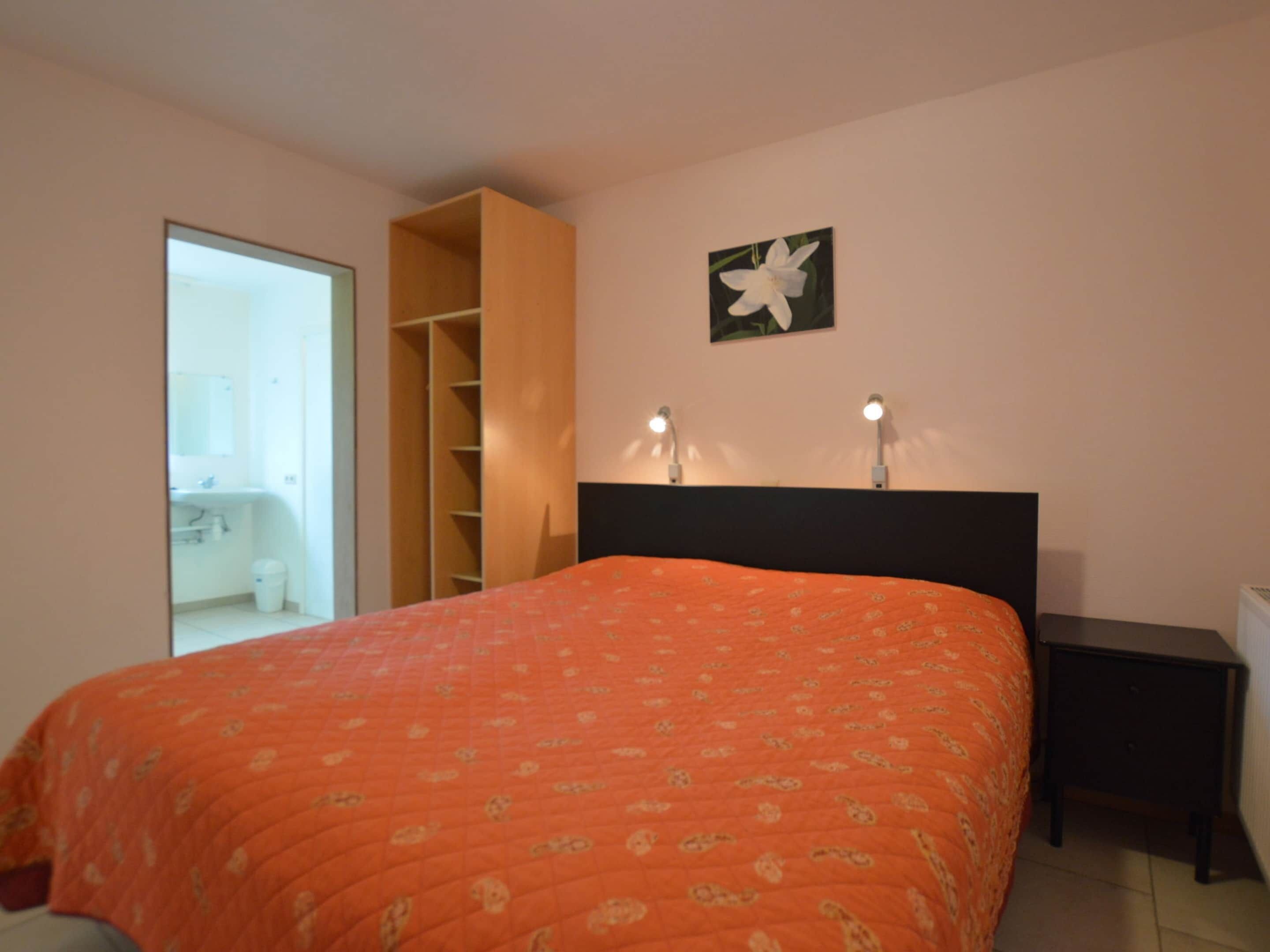 Quarto com cama, mesa de cabeceira, guarda-roupa e banheiro visível por uma porta aberta.
