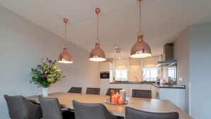 Uma sala de jantar moderna com uma mesa de madeira e cadeiras cinzentas, luminárias pendentes de cobre no teto e um vaso de flores sobre a mesa.