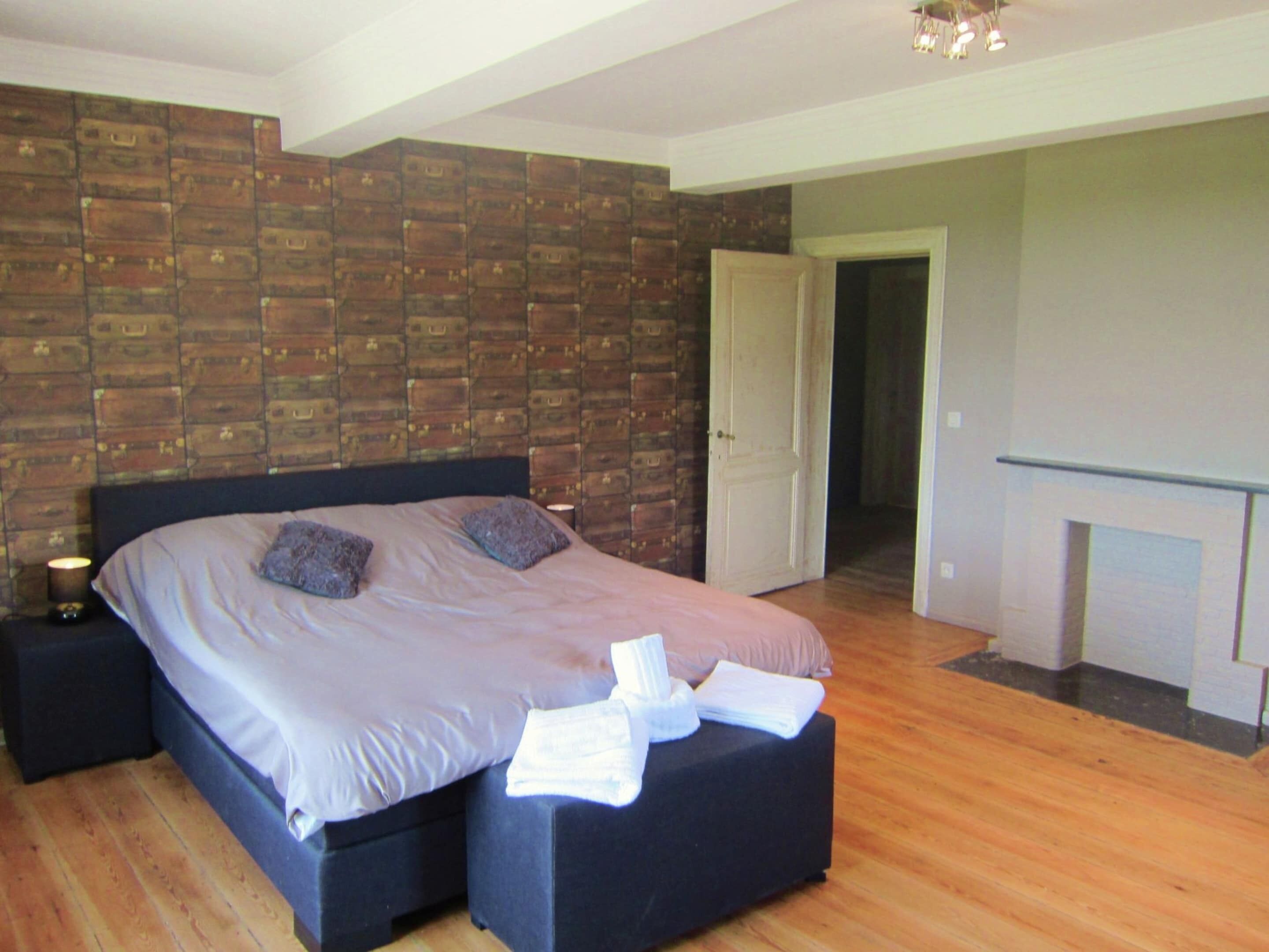 Quarto com uma cama grande, uma mesinha de cabeceira, parede com padrão de tijolos e uma porta que dá para outro cômodo.