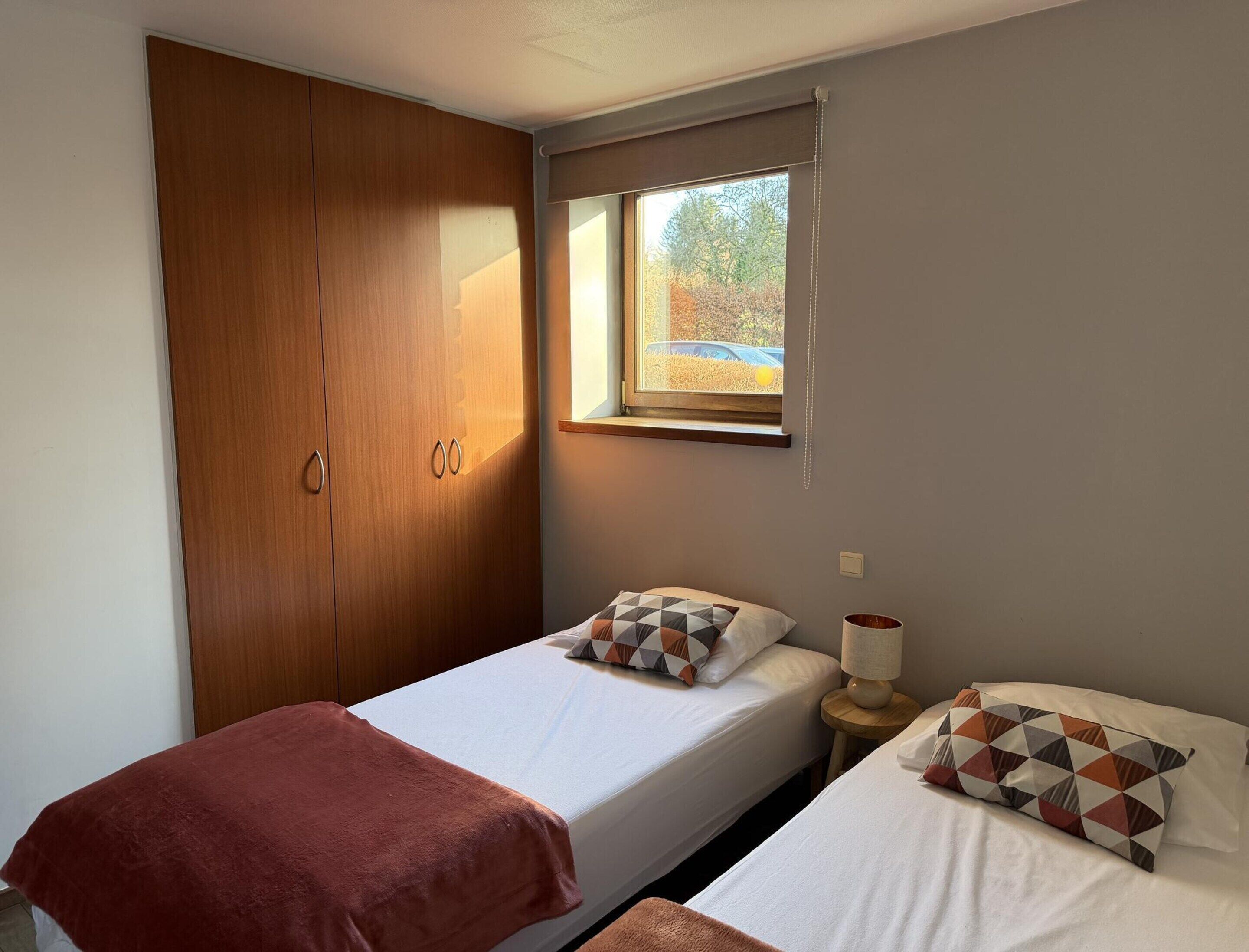 Quarto com duas camas de solteiro, guarda-roupa de madeira, janela com vista para o exterior e mesa de cabeceira com abajur.