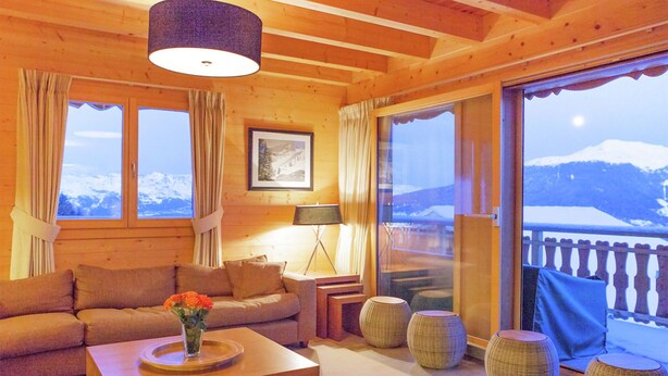 Interior acolhedor de uma cabana de madeira, com sofá, mesa de centro e vista para montanhas nevadas através de grandes janelas.