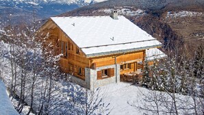 Uma cabana de madeira com telhado coberto de neve, rodeada por árvores e montanhas também cobertas de neve ao fundo.