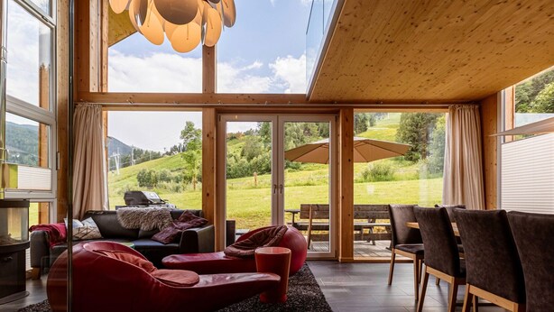Uma sala de estar ampla com um sofá vermelho, teto de madeira e janelas grandes que oferecem vista para uma paisagem verde.