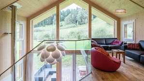 Uma sala de estar espaçosa com teto de madeira, grandes janelas com vista para uma paisagem verde, luminária pendente moderna e um puff vermelho.