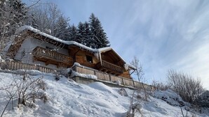 Um chalé de madeira com varanda, rodeado por árvores cobertas de neve e um céu limpo.