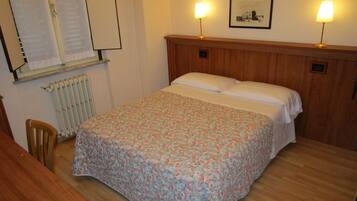 Quarto com cabeceira de madeira, duas luminárias de cabeceira, colcha florida, cadeira de madeira e radiador.