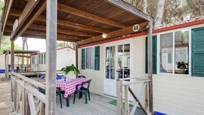 Um deck de madeira com mesa e cadeiras, uma área coberta com uma placa de sinalização e um prédio com persianas verdes.