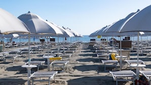 Uma praia com fileiras de espreguiçadeiras e guarda-sóis, alguns ocupados por pessoas.