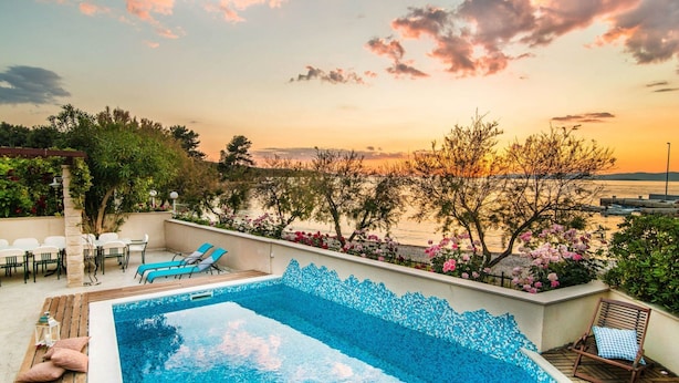 Piscina com azulejos azuis em mosaico, rodeada por espreguiçadeiras e um deck de madeira.