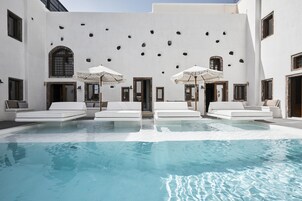 Área da piscina com cadeiras de sol brancas e guarda-sóis, piscina límpida e um edifício com fachada branca e janelas em arco.