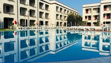 Piscina com cadeiras de descanso brancas e um edifício com várias varandas ao fundo.