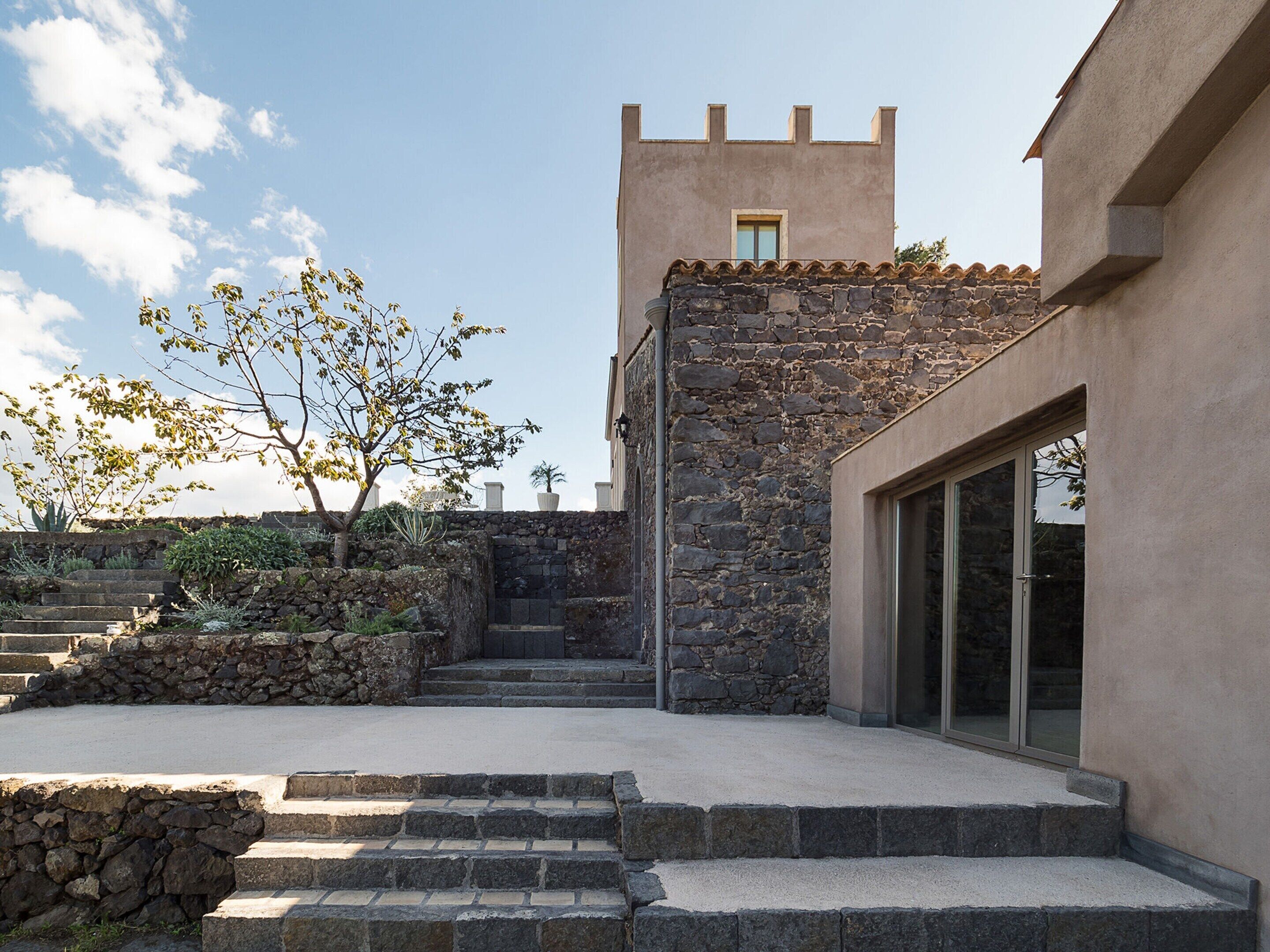 Um pátio com degraus de pedra que levam a uma porta, cercado por um muro de pedra e um edifício moderno com estrutura em torre.