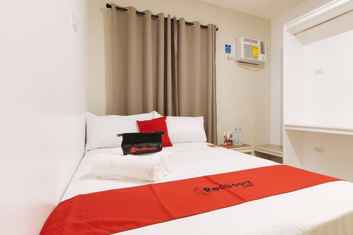 Quarto de hotel com uma cama, uma mala vermelha e preta, uma garrafa de água e um ar condicionado fixado na parede.