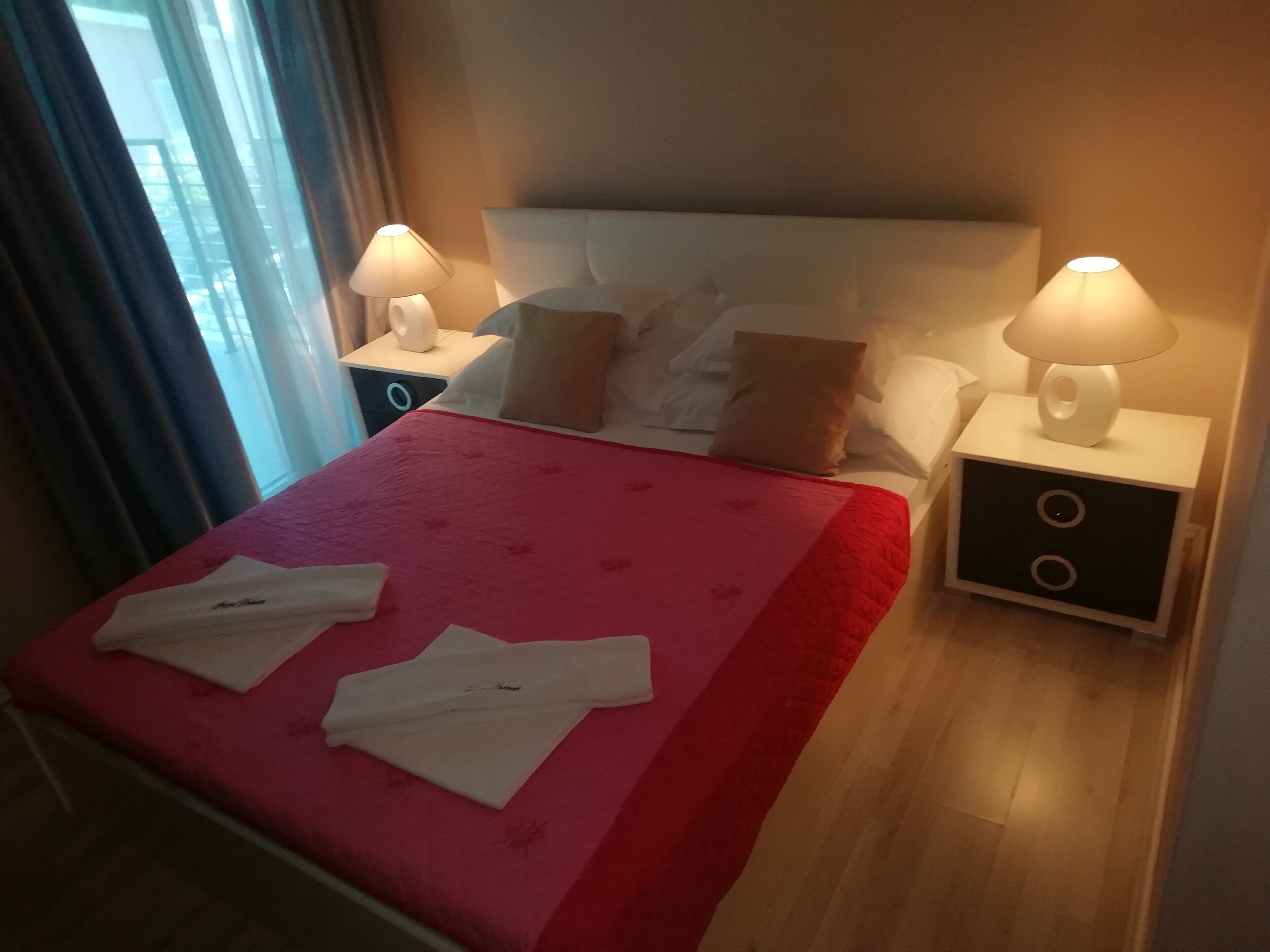 Quarto de hotel com colcha vermelha, duas almofadas, cabeceira branca, duas mesas de cabeceira com abajures e uma janela com cortinas.